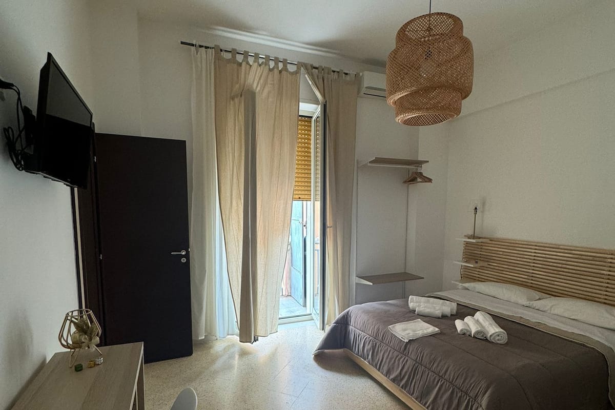 Quarto de hotel com cama, escrivaninha, cadeira, televisão e janela com cortinas.