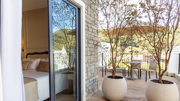 Quarto de hotel moderno com porta de vidro que dá para uma varanda, uma cama com roupa de cama bege e duas grandes plantas em vasos na varanda.