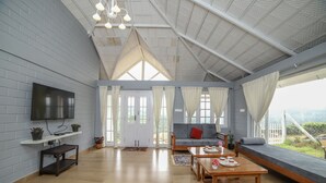 Uma sala de estar espaçosa, com teto alto, televisão de tela plana, sofá, mesa de centro de madeira e uma janela grande com cortinas.