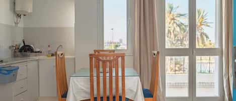 Uma sala de jantar com mesa e cadeiras, uma cozinha com pia e utensílios, e vista para palmeiras pela janela.