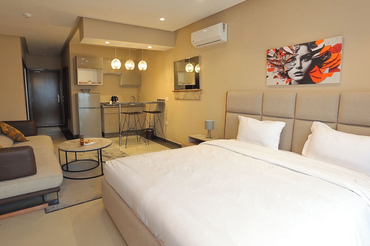 Quarto de hotel moderno com uma cama grande, um sofá, uma mesa redonda pequena e uma cozinha compacta.