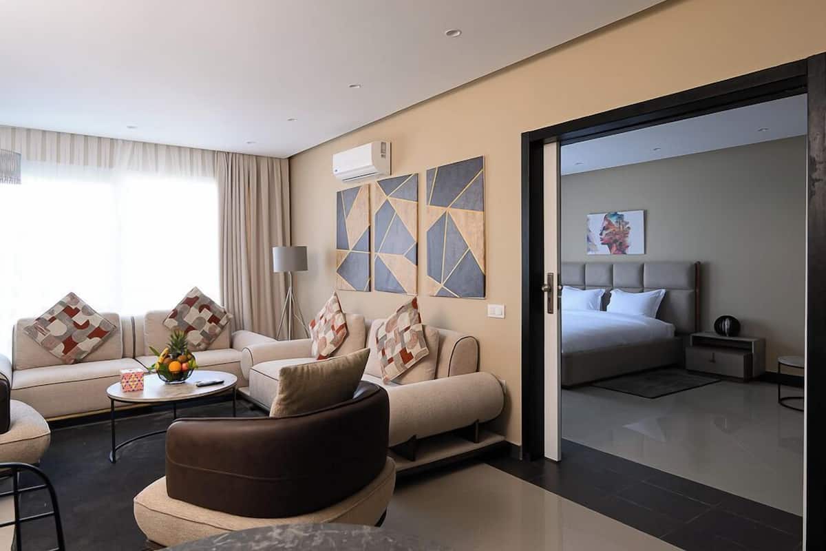 Quarto de hotel moderno com sofá, poltrona e cama. Há uma mesa de centro com uma fruteira e um abajur. O quarto tem uma janela grande com cortinas e obras de arte nas paredes.