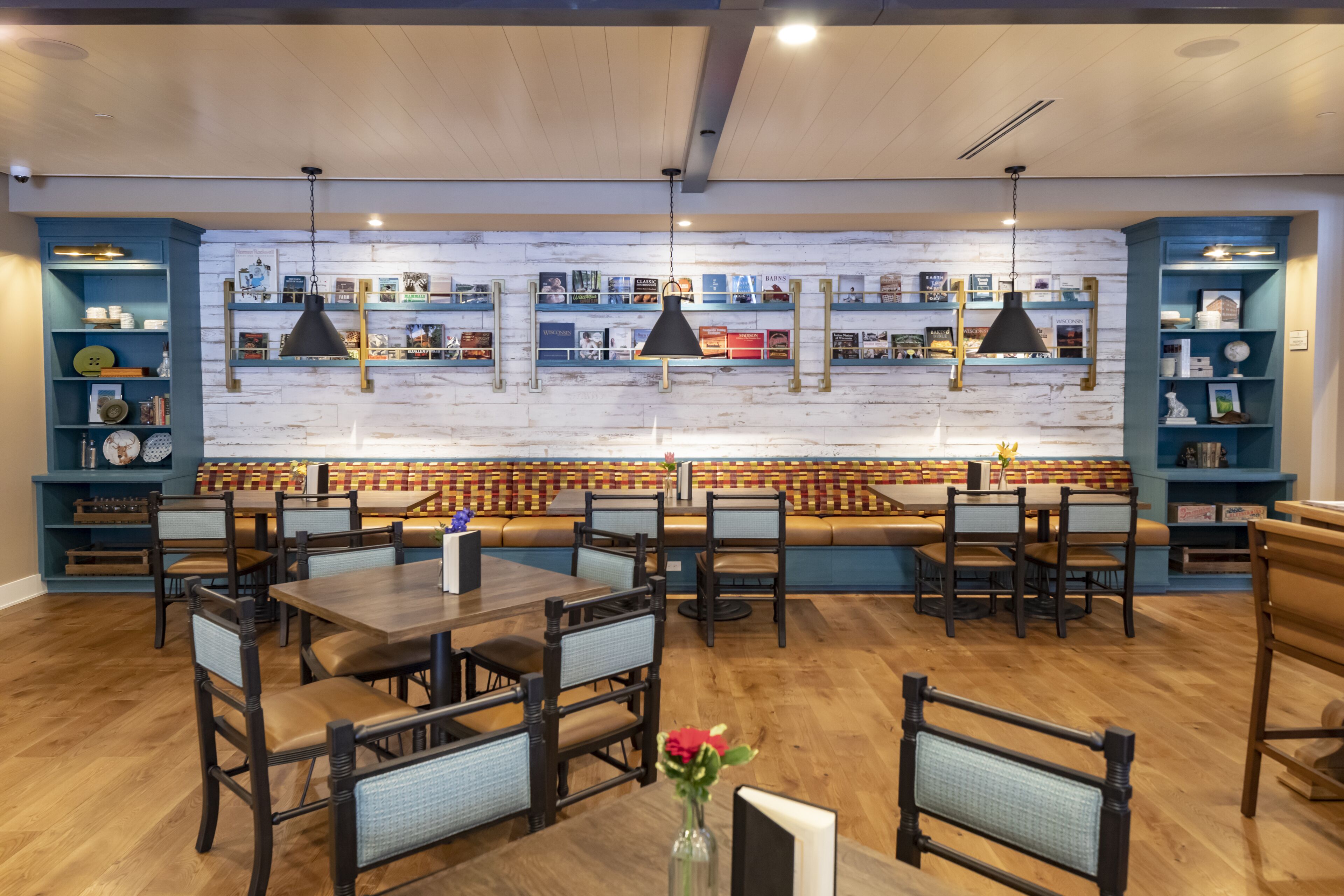 Um restaurante com mesas e cadeiras de madeira, uma parede de tijolos com quadros emoldurados e prateleiras azuis.