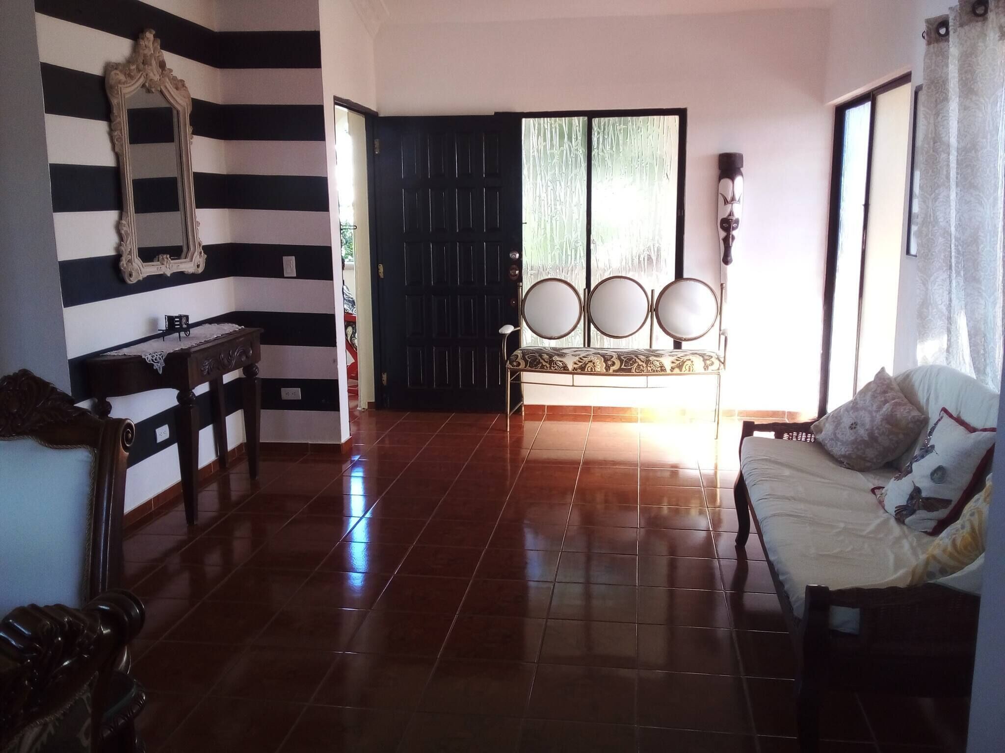 Um quarto com parede listrada, um espelho grande, um banco com espelhos redondos, um sofá com almofadas e uma porta com vista.