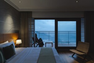 Quarto de hotel com cama, uma cadeira, uma mesa e varanda com vista para o mar.