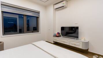 Quarto de hotel moderno com cama, televisão, mesa de cabeceira com produtos de higiene pessoal e janela com vista para a cidade.