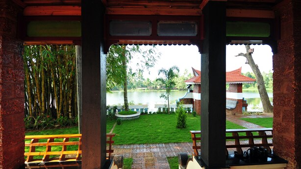 Área externa coberta com vista para um lago, um gramado e um edifício tradicional ao fundo.