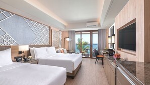 Quarto de hotel com duas camas, uma mesa com cadeira, televisão e varanda com vista para o mar.
