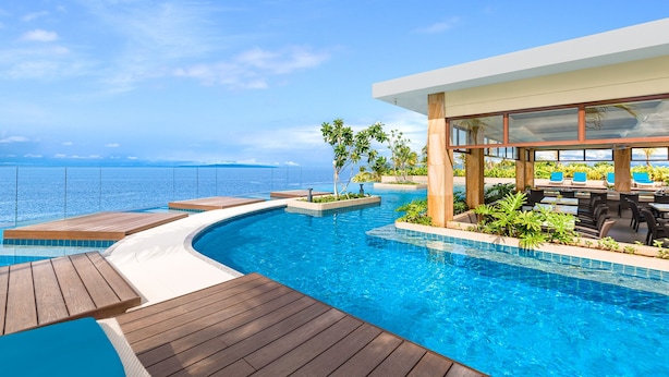 Uma piscina infinita moderna com um deck de madeira, rodeada por vegetação exuberante e vista para o oceano.