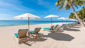 Uma praia com fileiras de espreguiçadeiras e guarda-sóis brancos, palmeiras e um céu azul límpido.