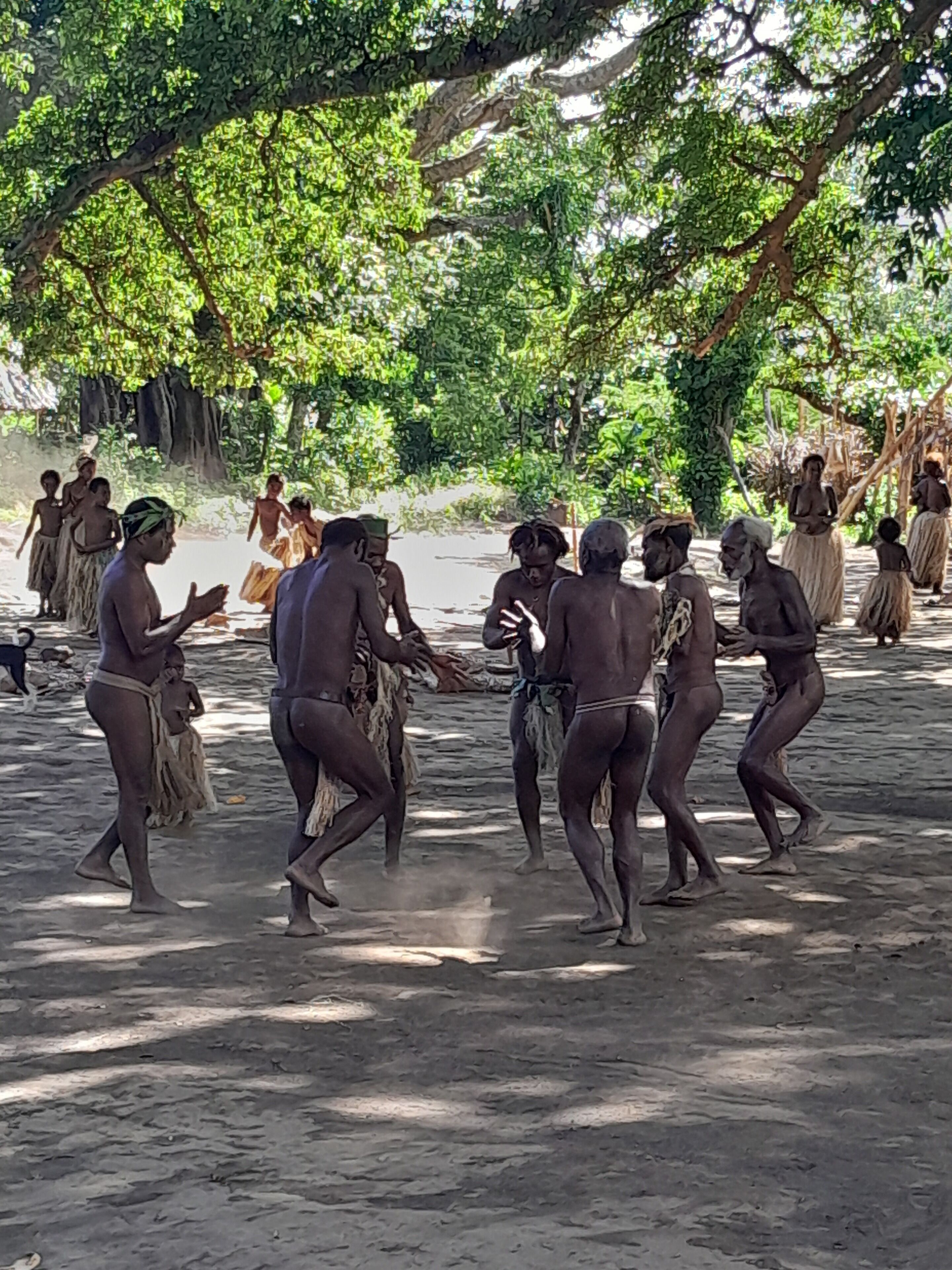 Um grupo de pessoas vestindo trajes tradicionais dançando em ambiente externo, com árvores e chão de terra.