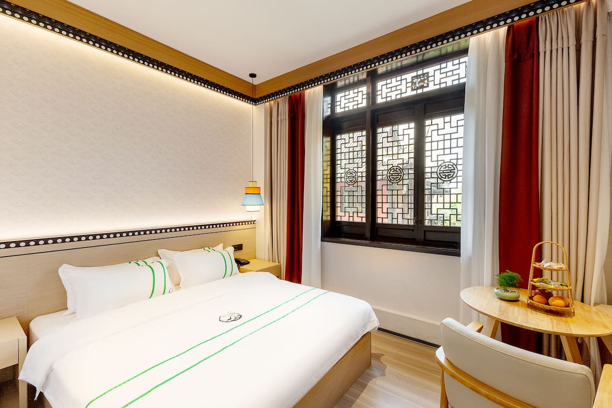 Quarto de hotel com uma cama grande, uma mesa de cabeceira, uma pequena mesa redonda com uma cesta de frutas e uma janela com vidros decorativos.