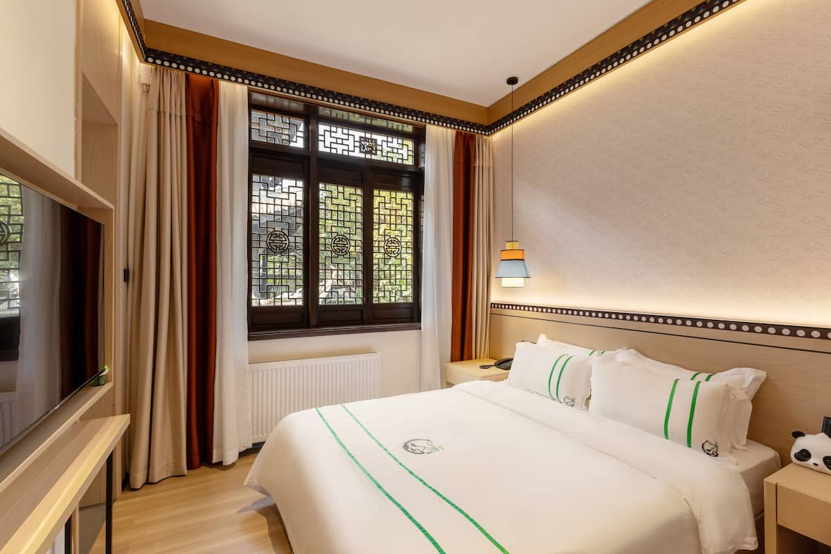 Quarto de hotel com cama grande, televisão, janela com cortinas e mesa de cabeceira com abajur.