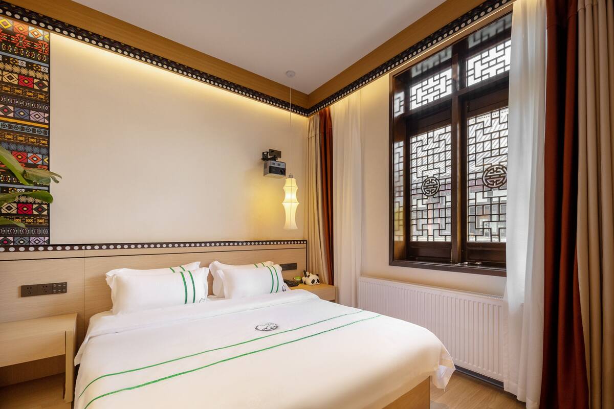 Quarto de hotel com cama bem arrumada, mesa de cabeceira com luminária, radiador e janela com painéis de vidro decorativos.