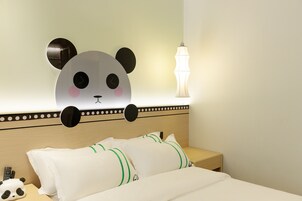 Cama com roupa de cama branca e listras verdes, mesa de cabeceira com abajur e decoração de panda fixada na parede.