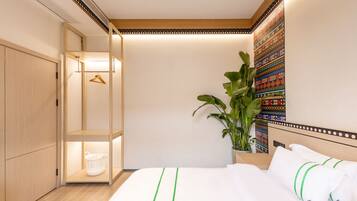 Quarto de hotel com cama, guarda-roupa, cabideiro, planta e parede com padrão decorativo.