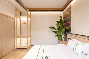 Quarto de hotel com cama, guarda-roupa, cabideiro, planta e parede com padrão decorativo.