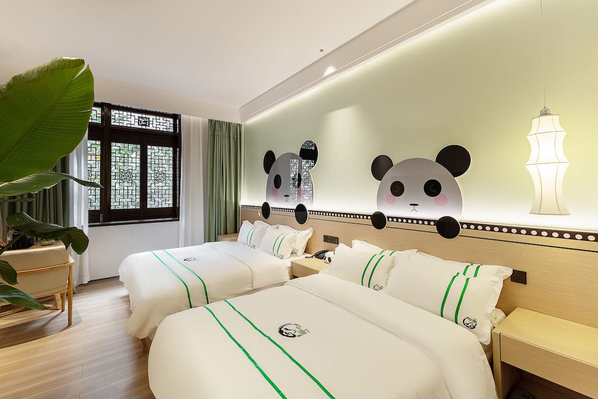 Quarto de hotel com duas camas, uma planta grande, janela com cortinas e decorações nas paredes.