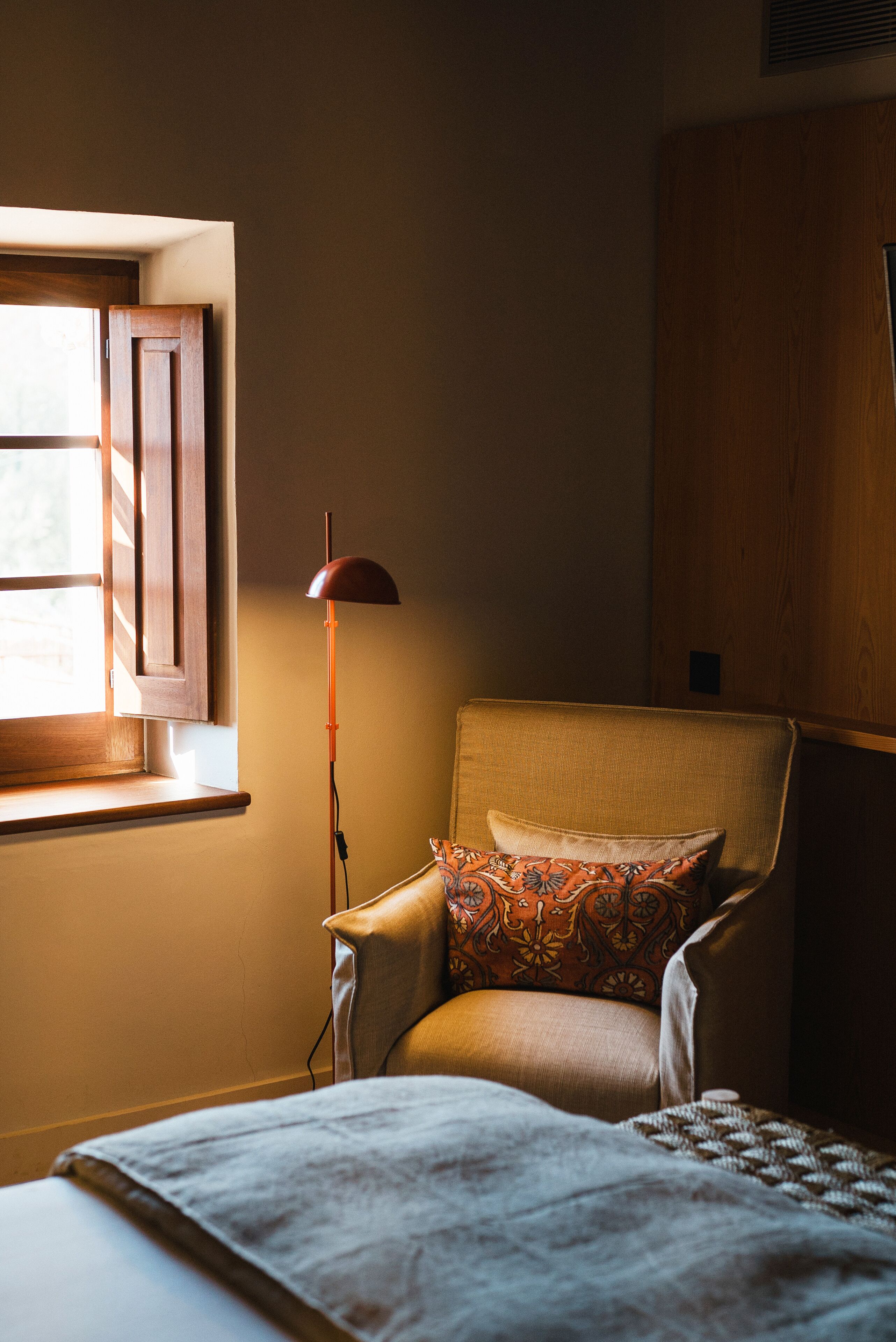 Um quarto com um armário de madeira, uma cadeira com almofada estampada, um abajur e uma cama com manta.