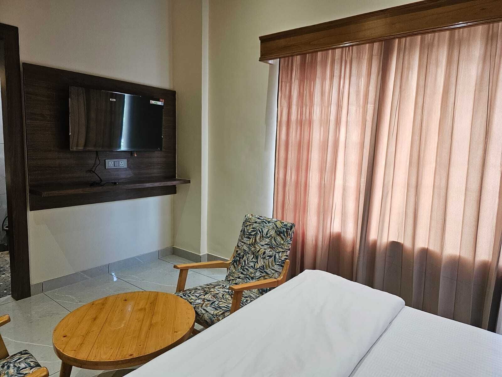 Quarto de hotel com uma cama, uma televisão presa a um painel de madeira escura, uma poltrona estampada, uma mesa de madeira redonda e uma janela com cortinas.