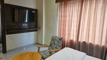 Quarto de hotel com uma cama, uma televisão presa a um painel de madeira escura, uma poltrona estampada, uma mesa de madeira redonda e uma janela com cortinas.