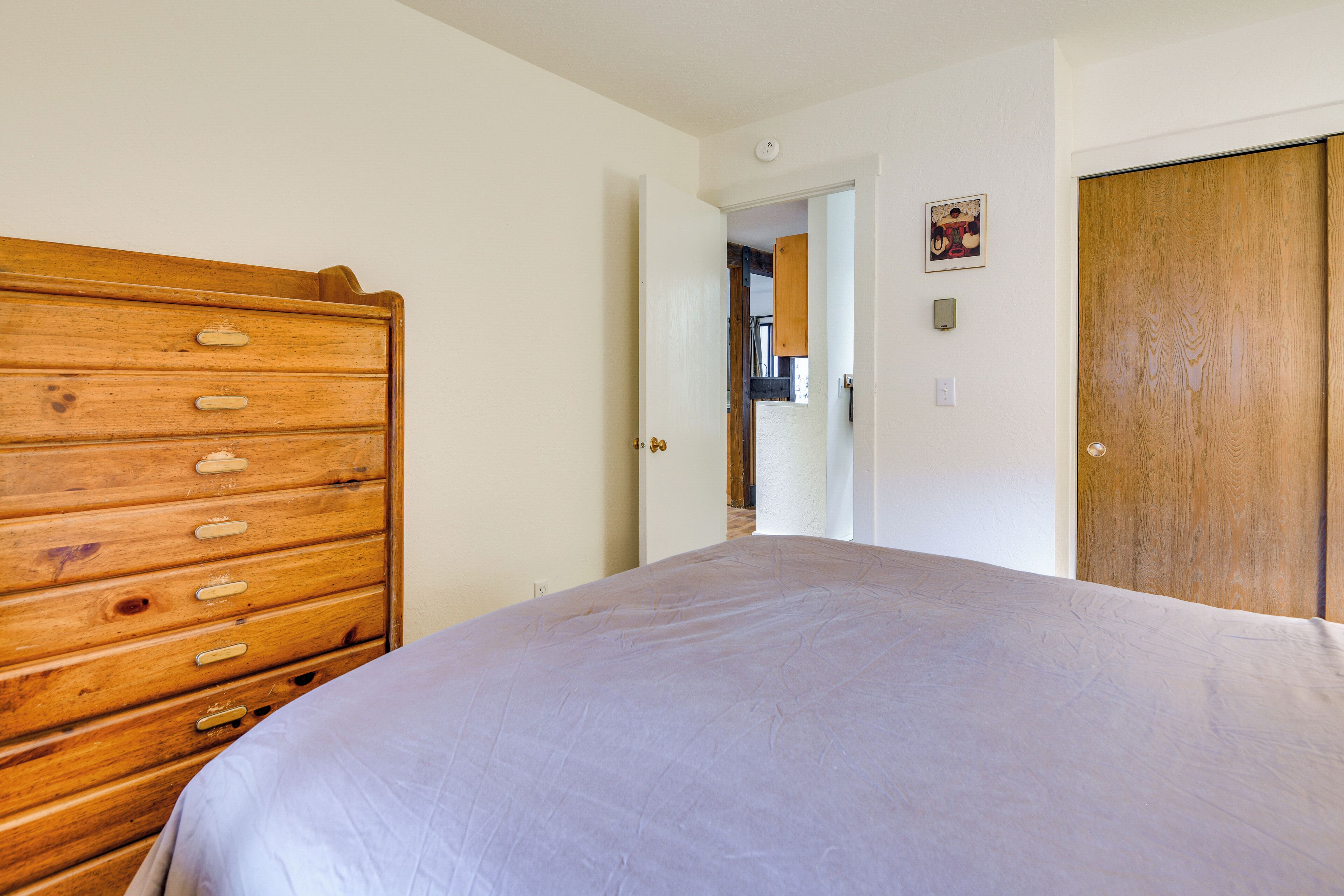 Quarto com cômoda de madeira, cama com colcha clara, porta de madeira e corredor que dá para outro quarto.