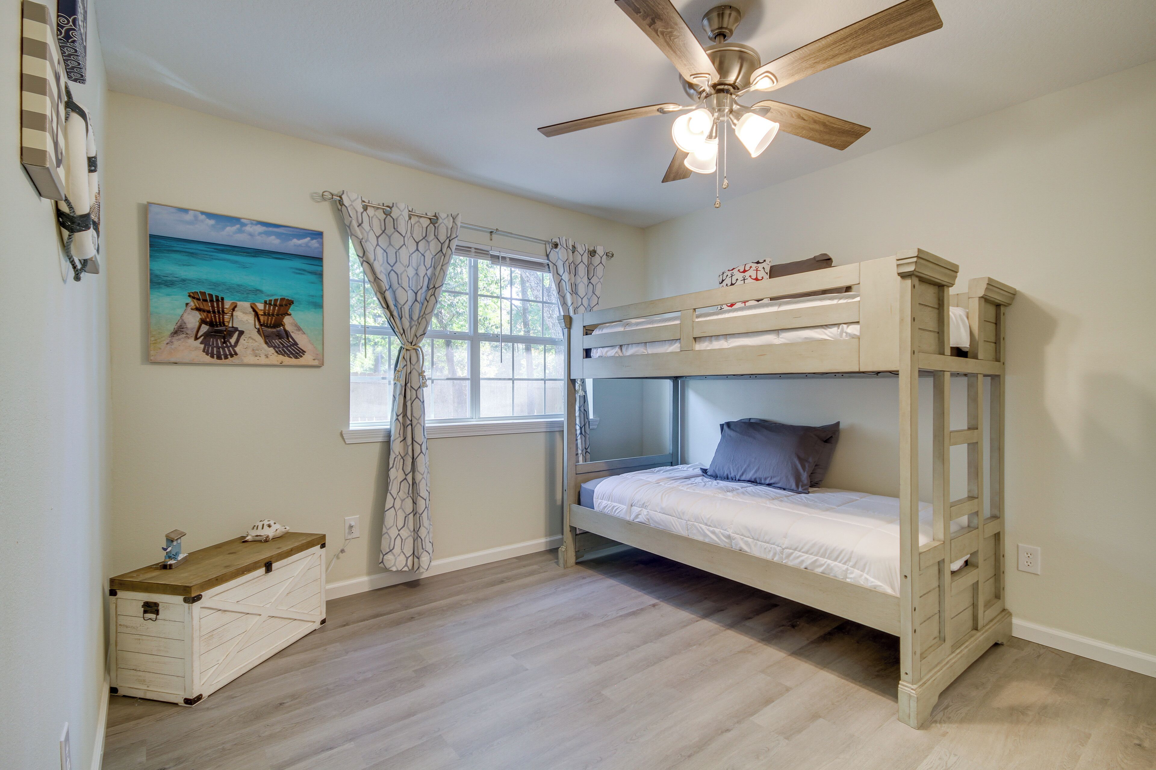 Quarto com beliche, ventoinha no teto, janela com cortinas, um quadro e cômoda de madeira.