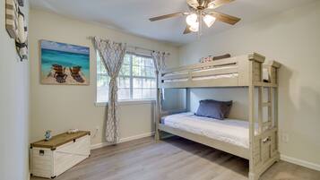 Quarto com beliche, ventoinha no teto, janela com cortinas, um quadro e cômoda de madeira.