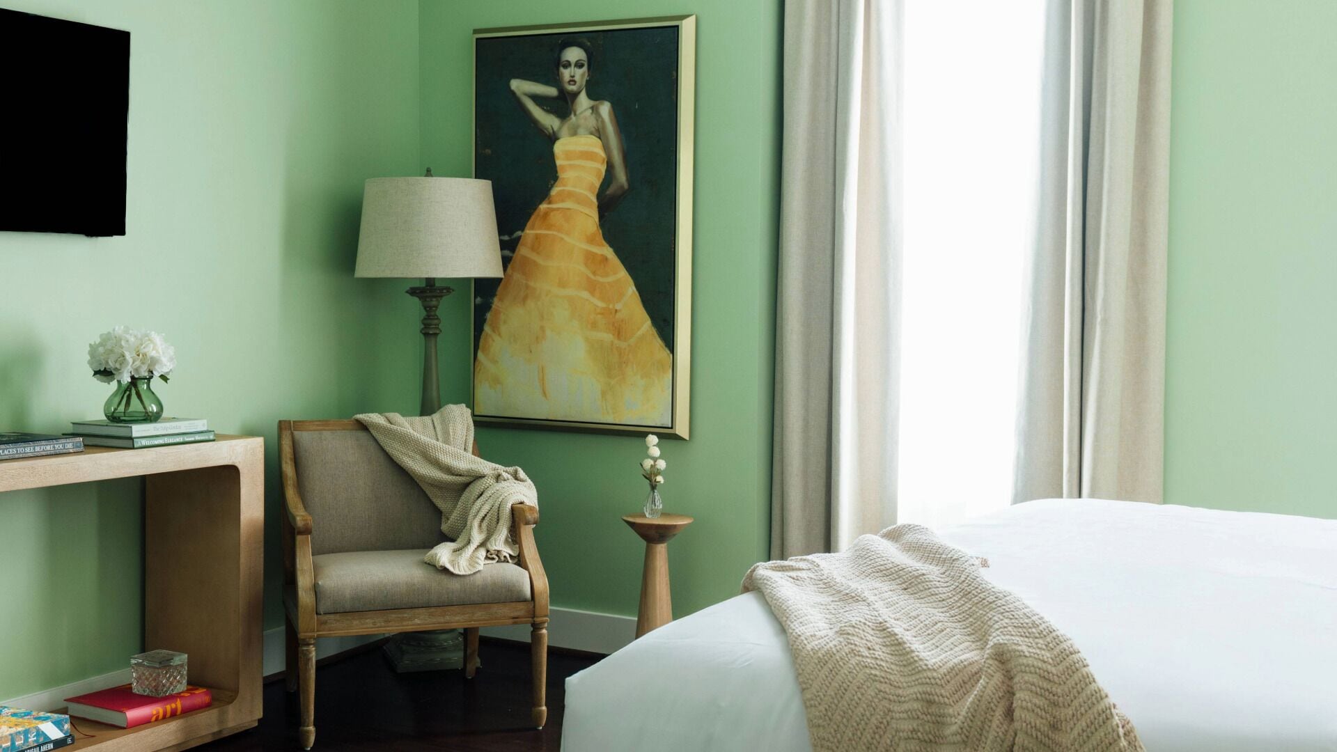 Quarto de hotel com uma cama, uma cadeira, uma escrivaninha com luminária, um quadro e um vaso com flores.