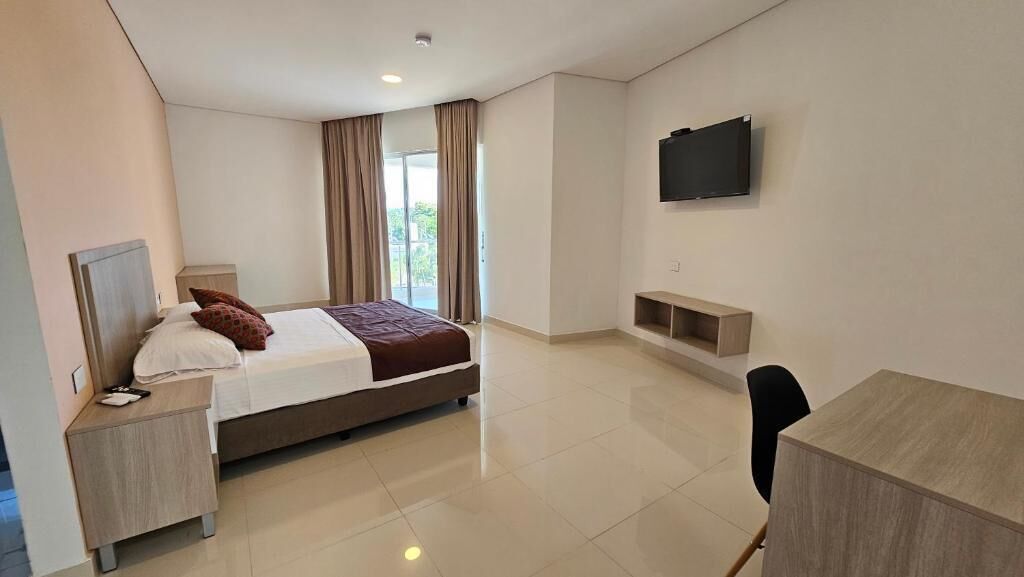 Quarto de hotel moderno com cama grande, escrivaninha, cadeira, TV fixada na parede e varanda com cortinas.