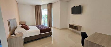 Quarto de hotel moderno com cama grande, escrivaninha, cadeira, TV fixada na parede e varanda com cortinas.