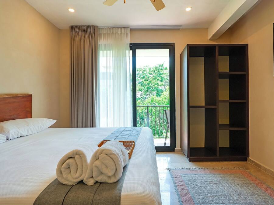 Quarto de hotel com cama, uma janela grande com cortinas, televisão de tela plana na parede e varanda com vista para áreas verdes.