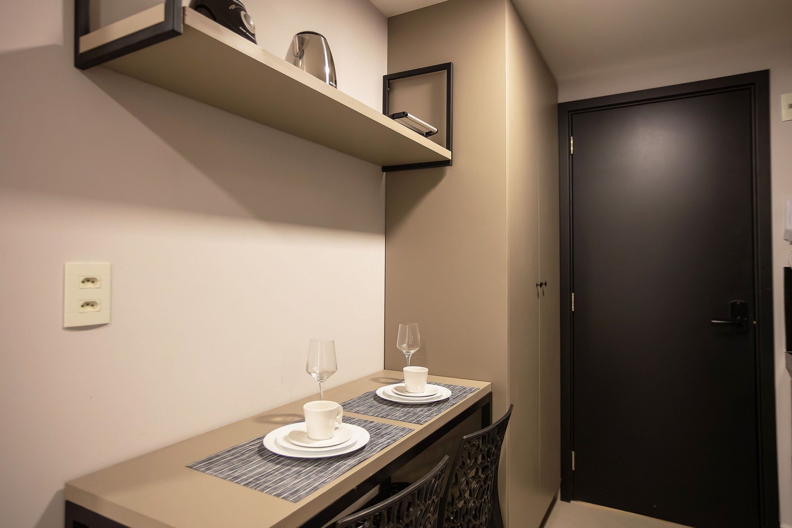 Quarto de hotel com uma mesa de jantar posta para dois, uma porta preta e uma prateleira acima da mesa.