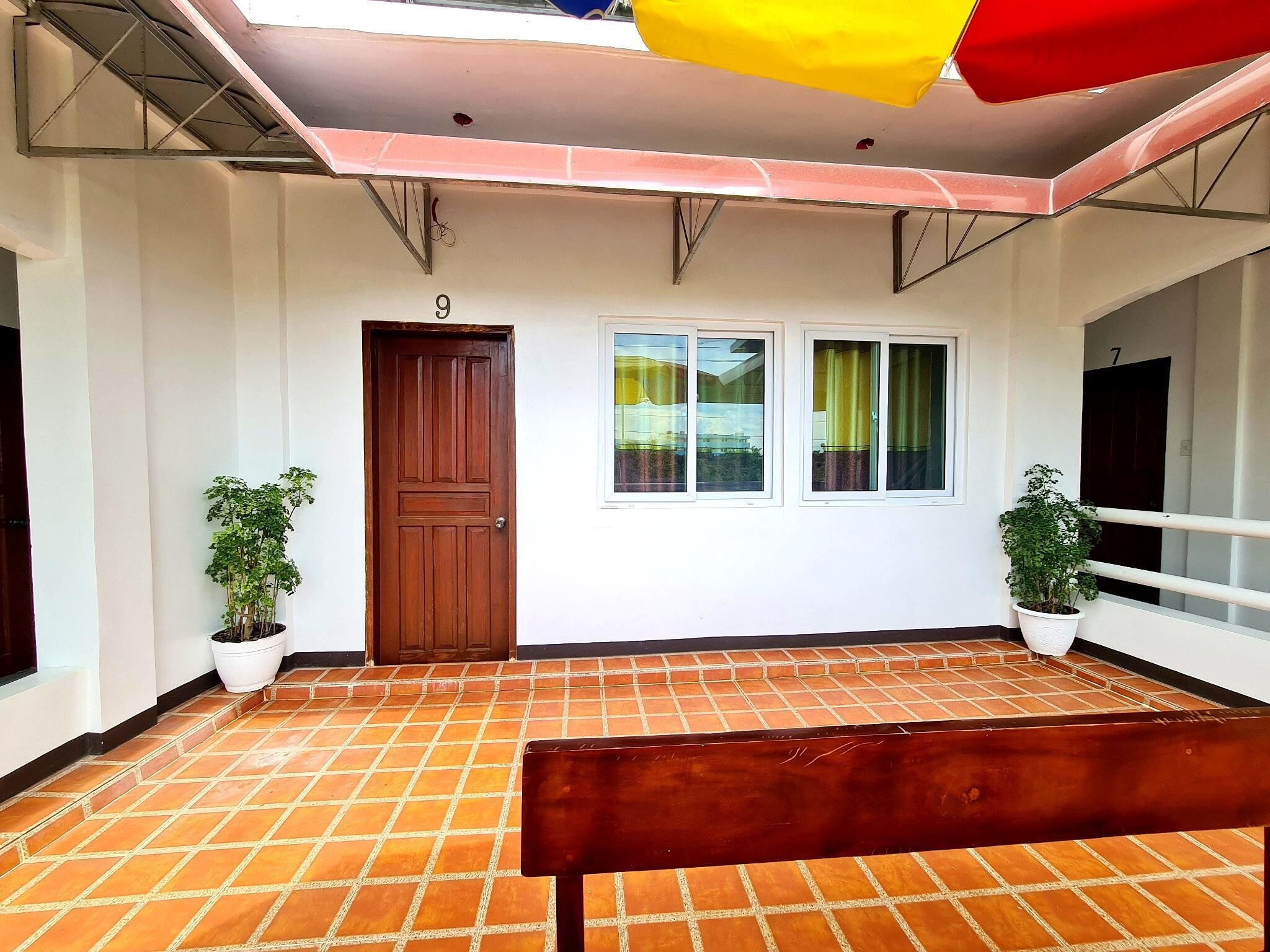 Uma área externa coberta com piso de azulejos, um banco de madeira, duas plantas em vasos e uma porta com o número 9.