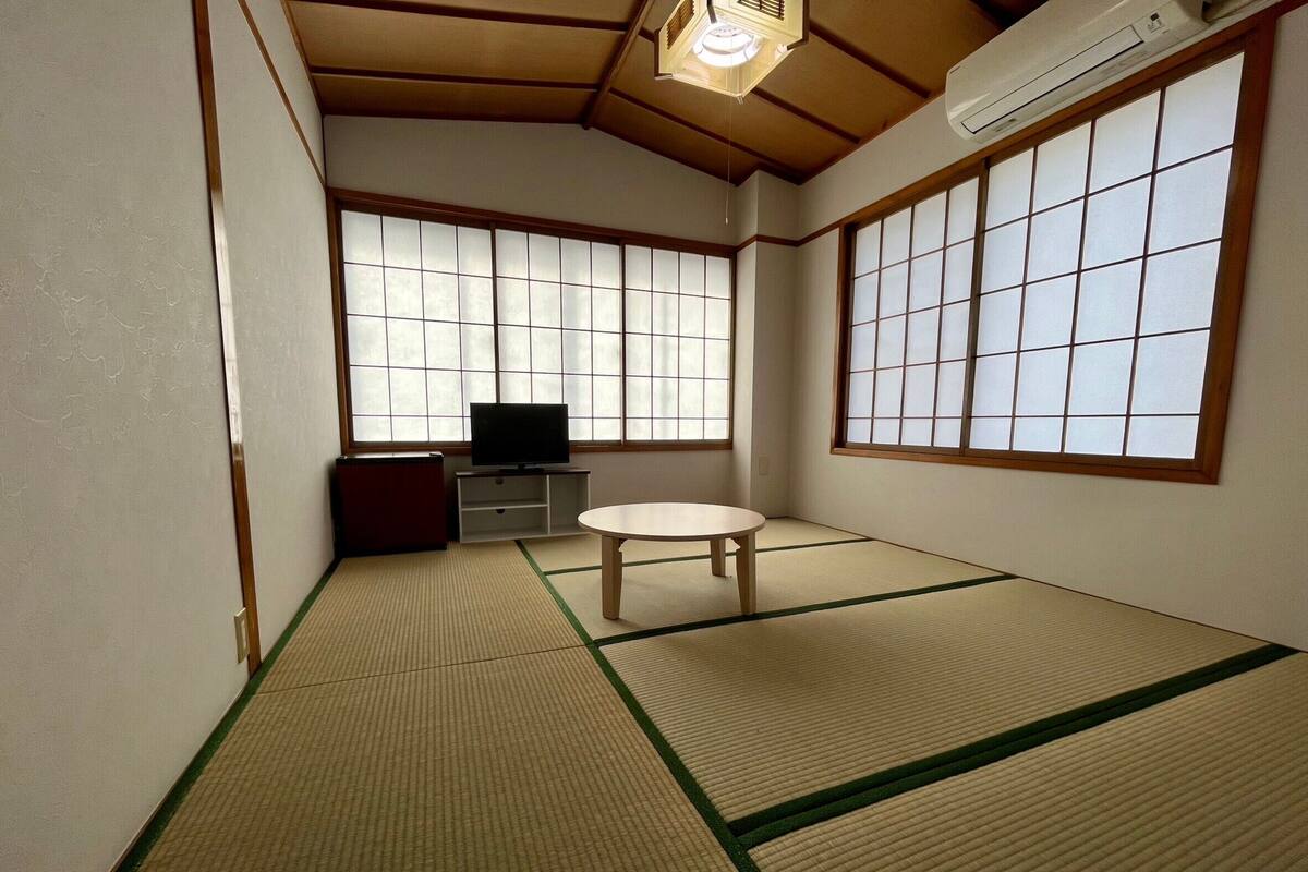 Um quarto japonês tradicional com piso de tatame, uma mesa de tatame e uma televisão sobre um suporte baixo, além de amplas divisórias shoji.