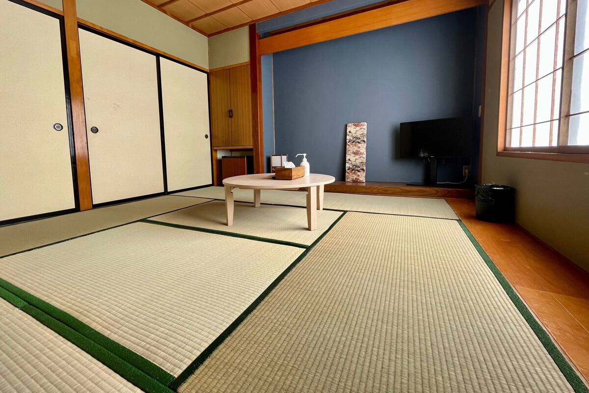 Um quarto tradicional japonês com esteiras de tatame, uma mesa baixa, uma televisão e portas deslizantes.