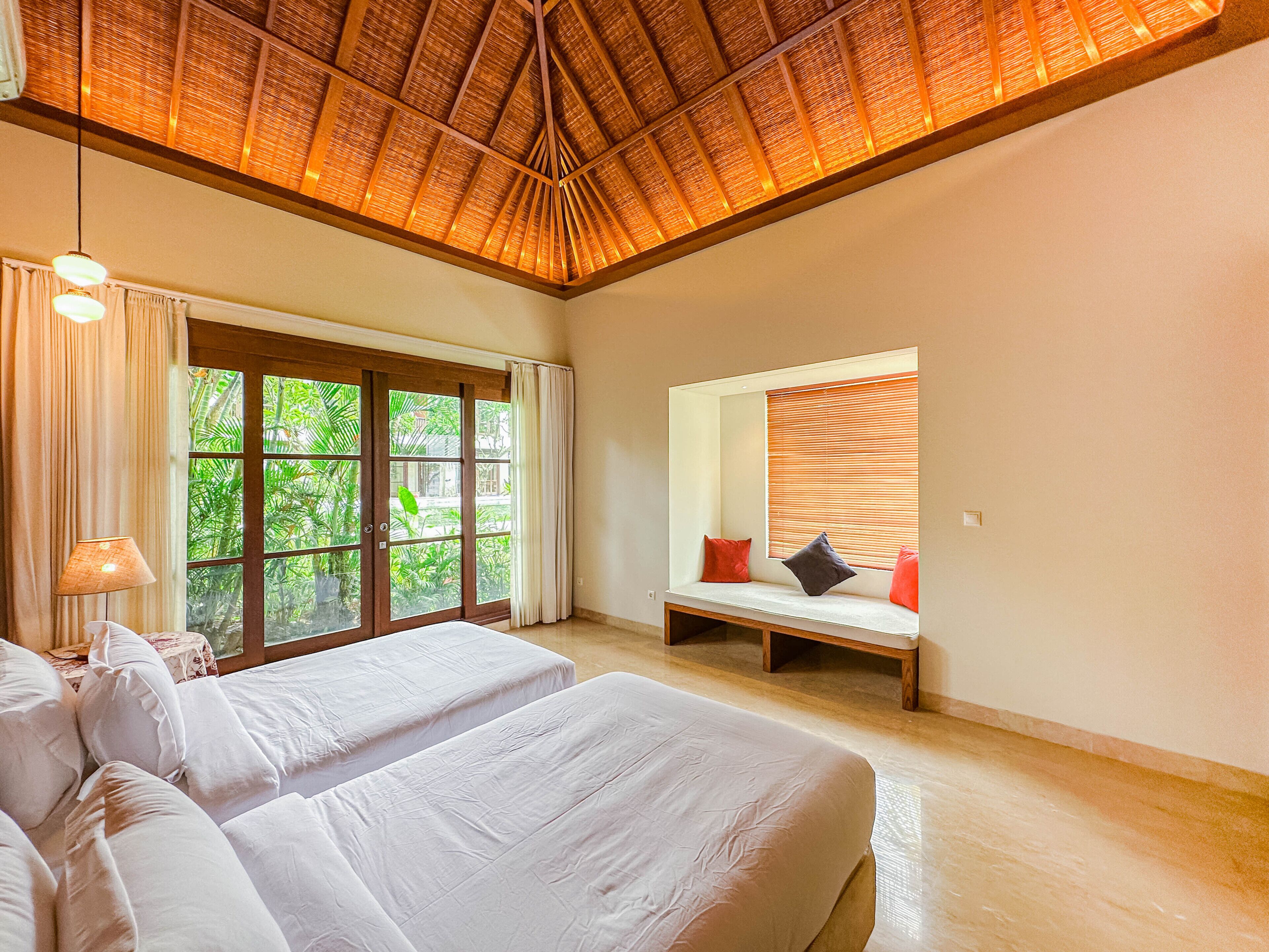 Quarto com duas camas, uma janela grande com cortinas, teto com detalhe em tecido e um banco com almofadas.