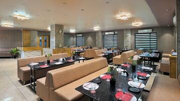 Ampla sala de jantar de hotel, com várias mesas e cabines, equipada com iluminação moderna e uma grande janela que proporciona luz natural.