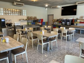 Um café moderno com várias mesas e cadeiras, um espaço para café e uma televisão fixada na parede.