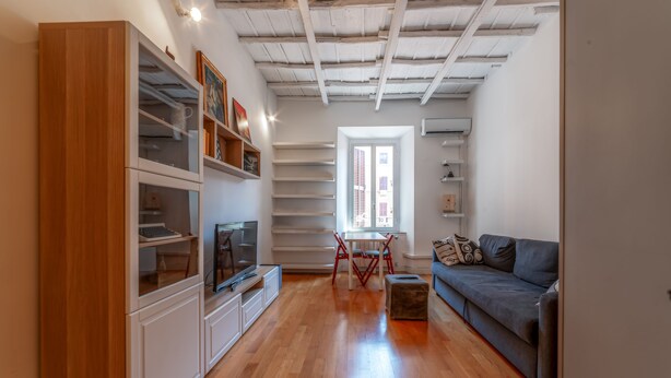 Uma sala de estar moderna com piso de madeira, um sofá cinzento, uma mesa de jantar com cadeiras e estantes embutidas com uma televisão.