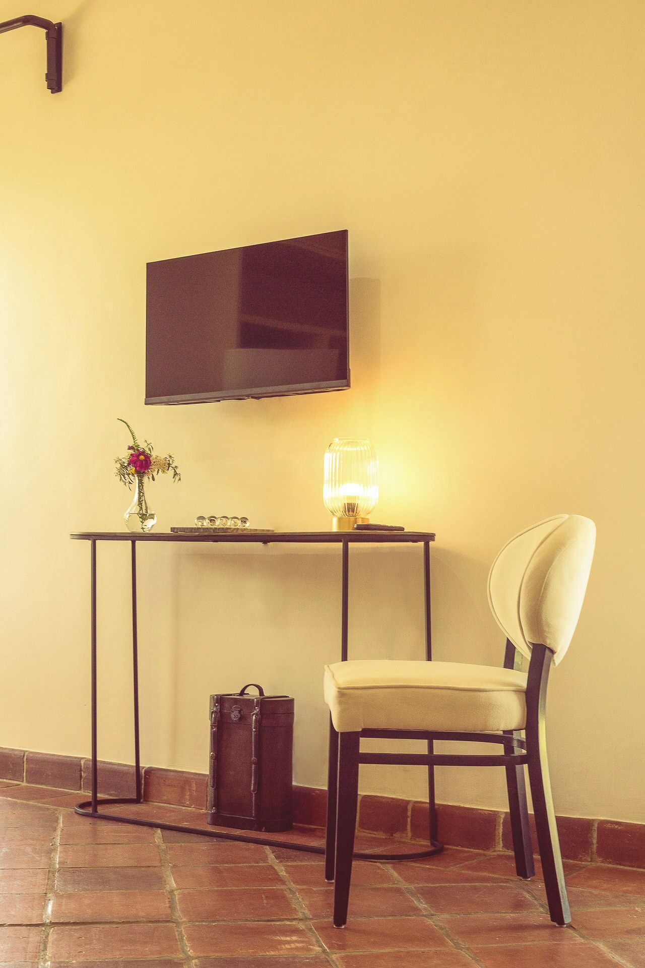 Um quarto pequeno com escrivaninha, cadeira, televisão, abajur, vaso com flores e mala.