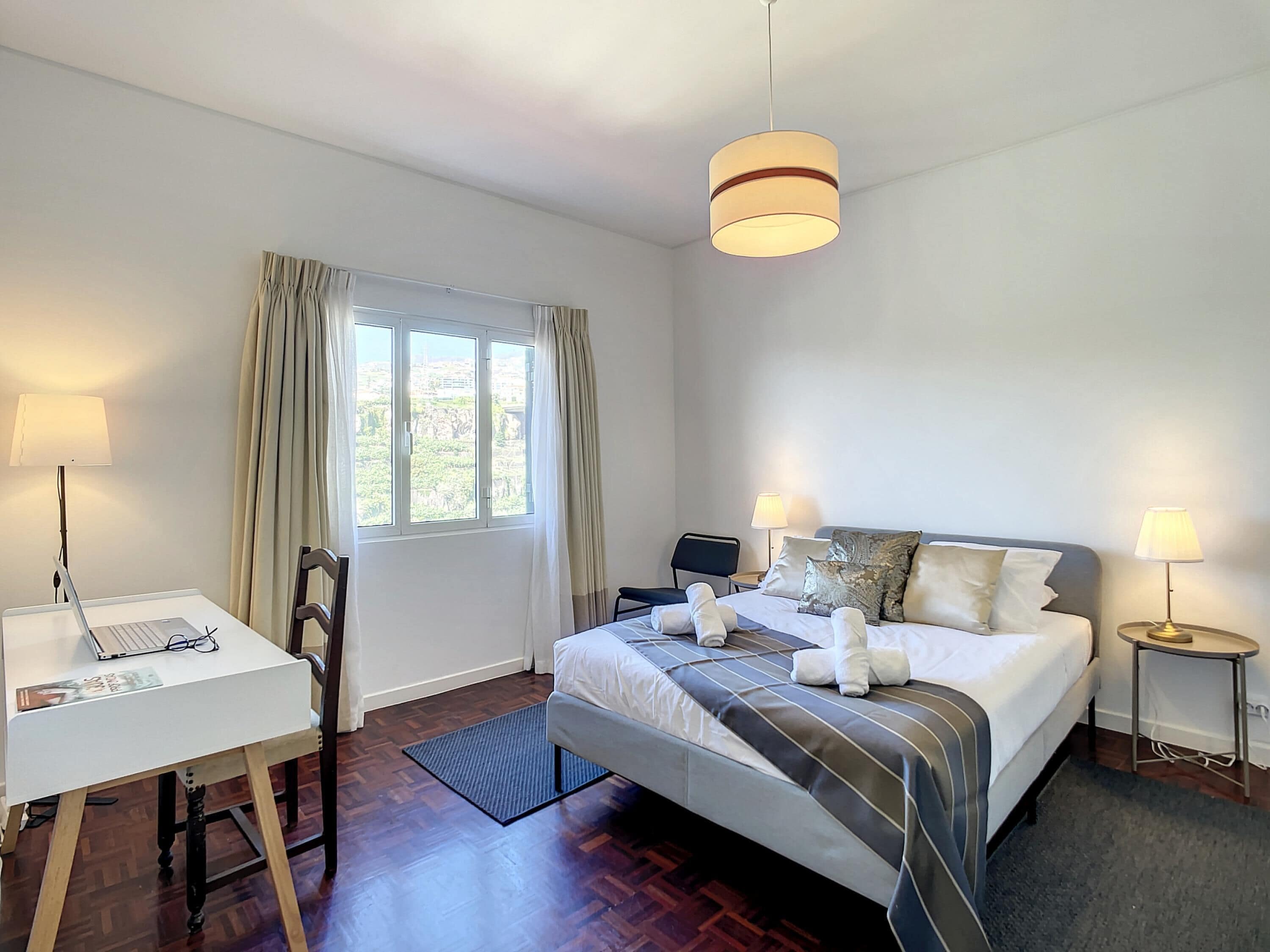Quarto de hotel com cama, escrivaninha, cadeira e janela com cortinas.