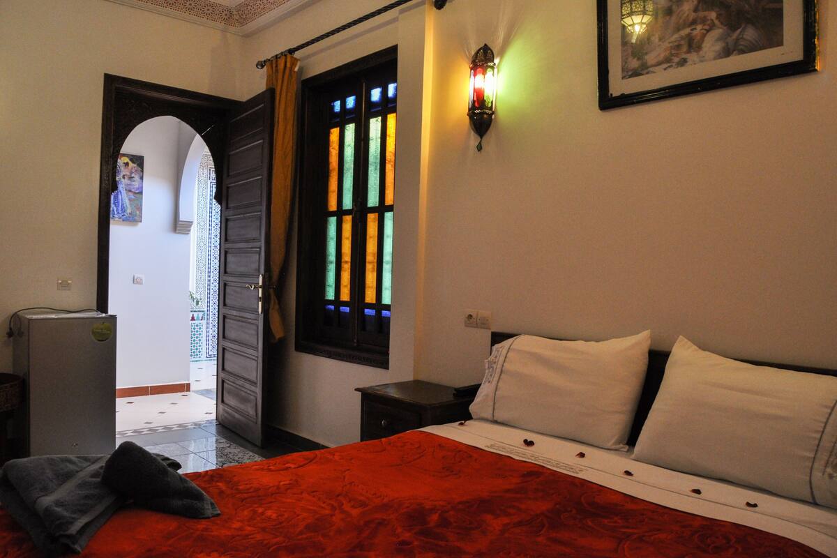 Quarto de hotel com cama, mesa de cabeceira, luminária na parede, porta com vista e geladeira.
