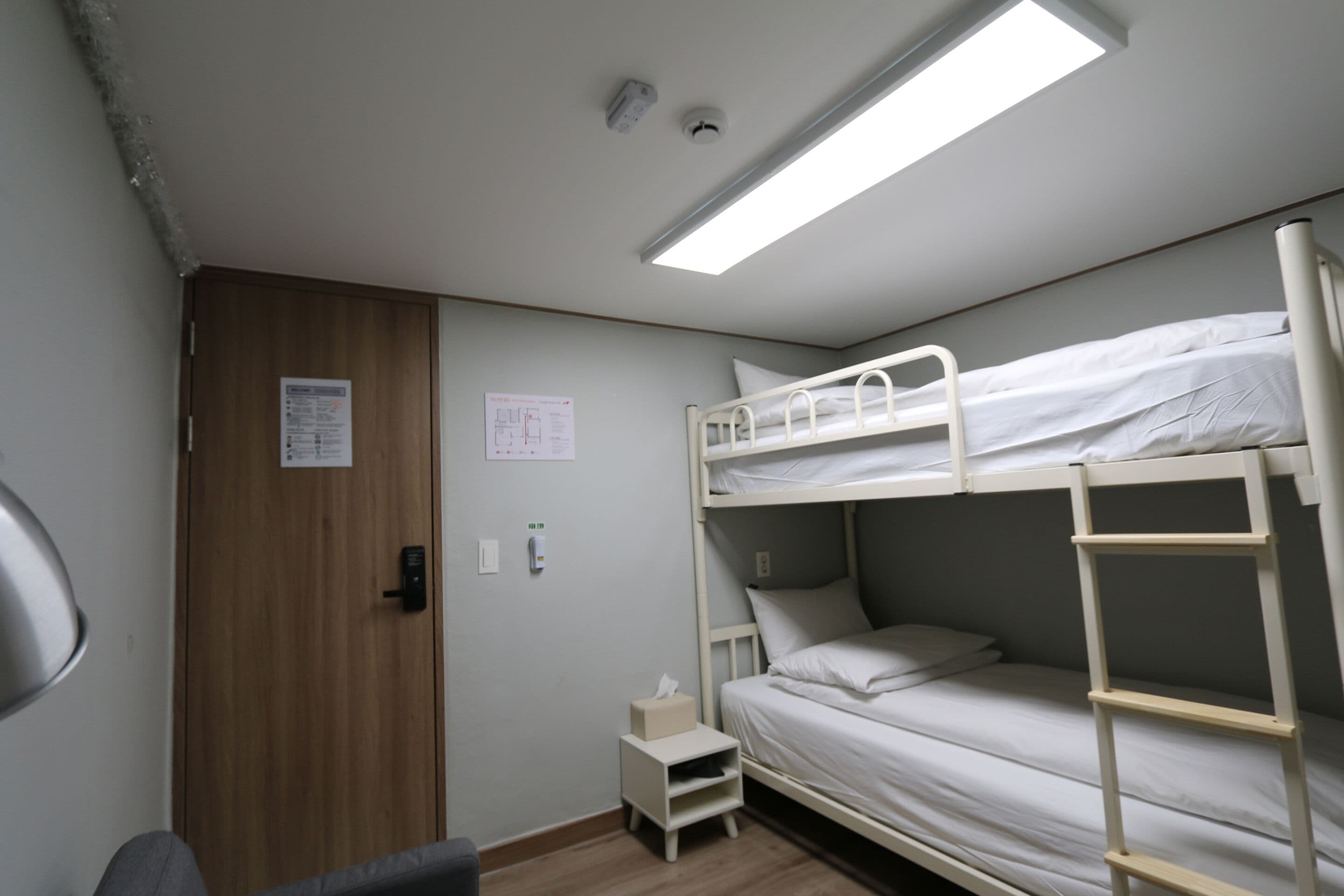 Quarto com beliche, porta de madeira, uma pequena mesa de cabeceira e luz no teto.