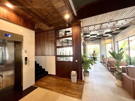 Um saguão de hotel com escada de madeira, balcão de recepção e área de estar com plantas em vasos.