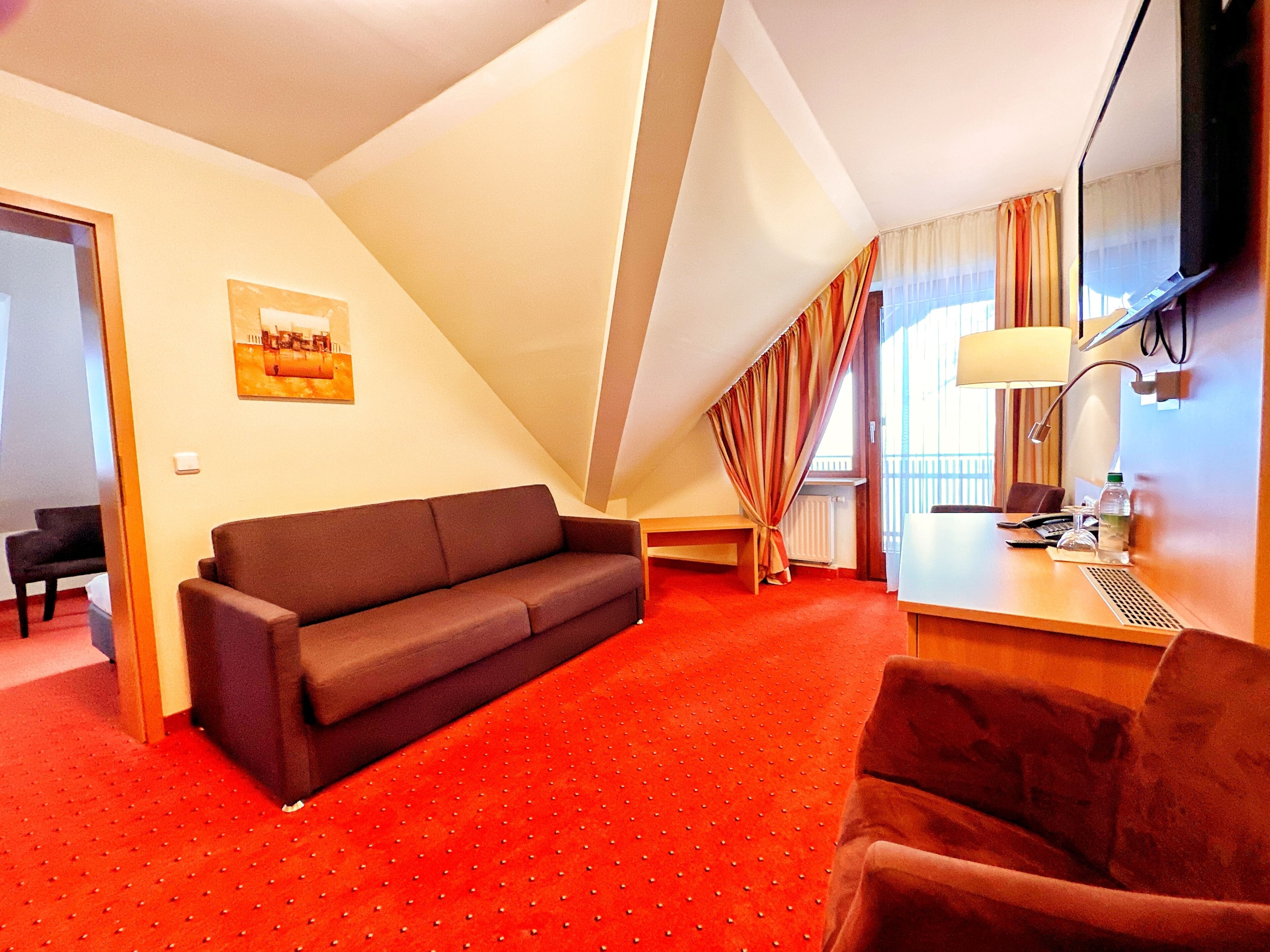 Quarto de hotel com um sofá marrom, uma escrivaninha de madeira, uma luminária e uma televisão.