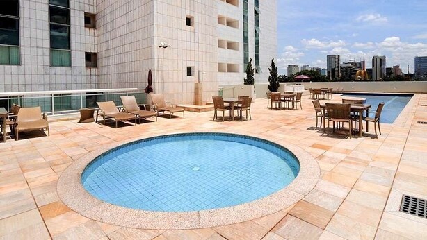 Área de piscina no terraço, com espreguiçadeiras e mesas, cercada por um edifício alto.