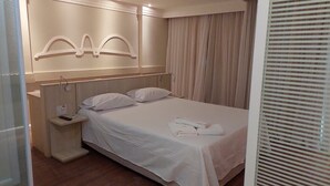 Quarto de hotel com cama, mesa de cabeceira, luminária na parede e janela com cortinas.