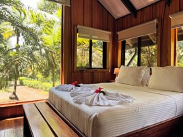 Cama com roupa de cama branca e uma flor vermelha, paredes de madeira e vista para o verde lá fora.