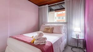 Quarto de hotel com cama, janela com cortinas, mesa de cabeceira com abajur e vista para um edifício.